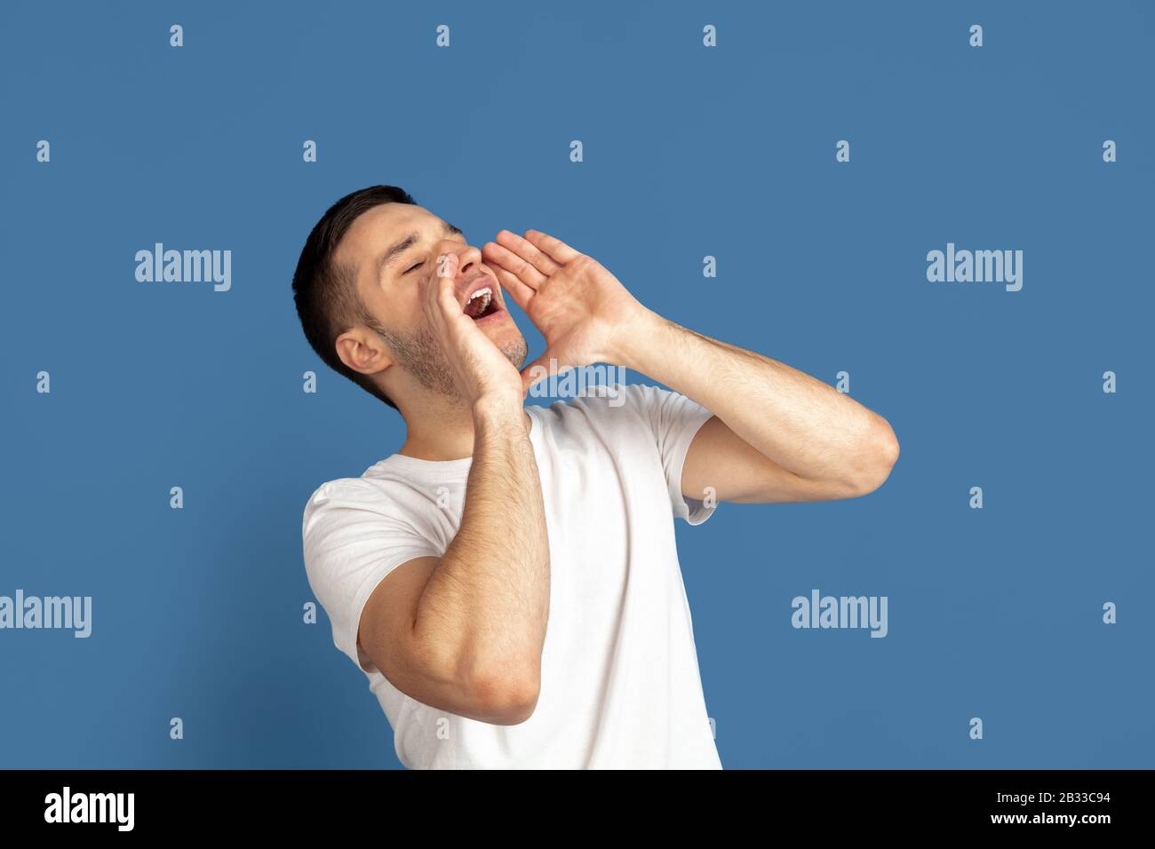 Calling, shouting. Caucasian young man's portrait on blue studio ...