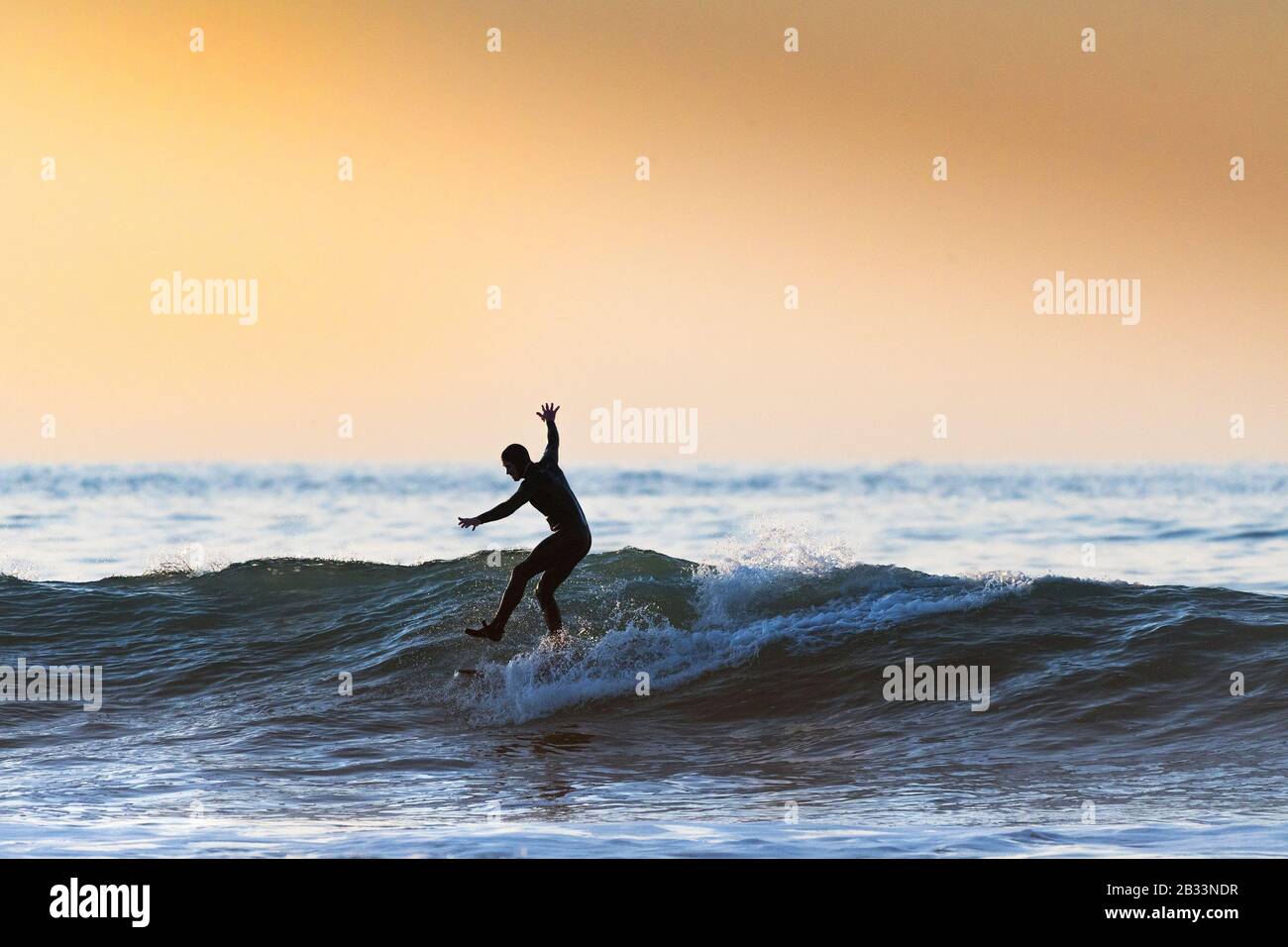 A surfer riding a wave and standing on one leg in evening light at ...