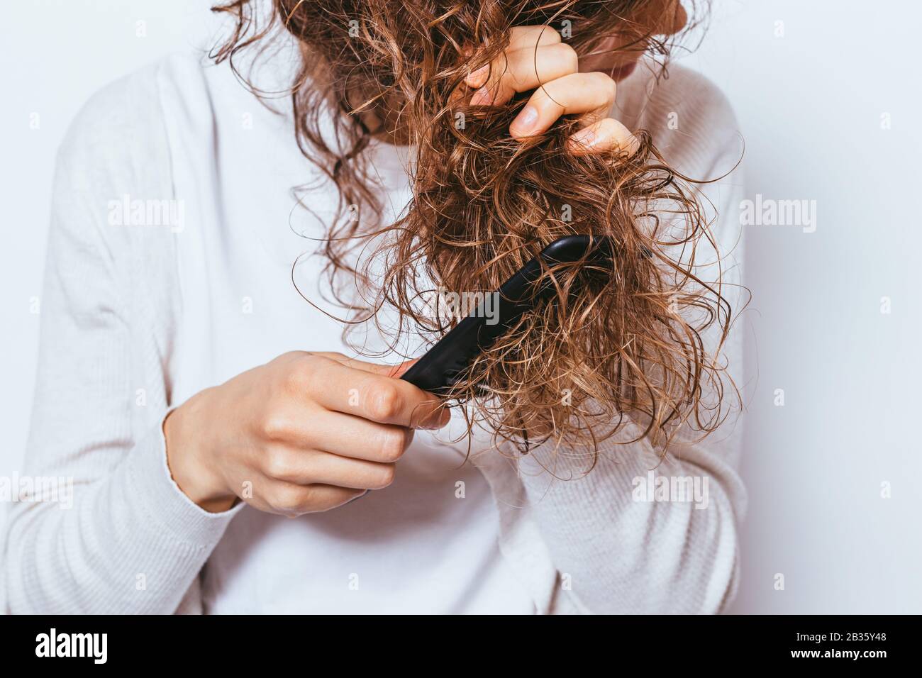 Close-up young woman wearing white shirt combing her tangled curly hair ...