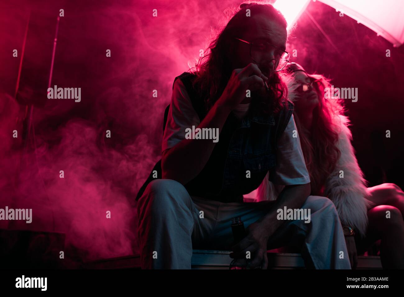 man smoking cigarette near young woman during rave party in nightclub ...