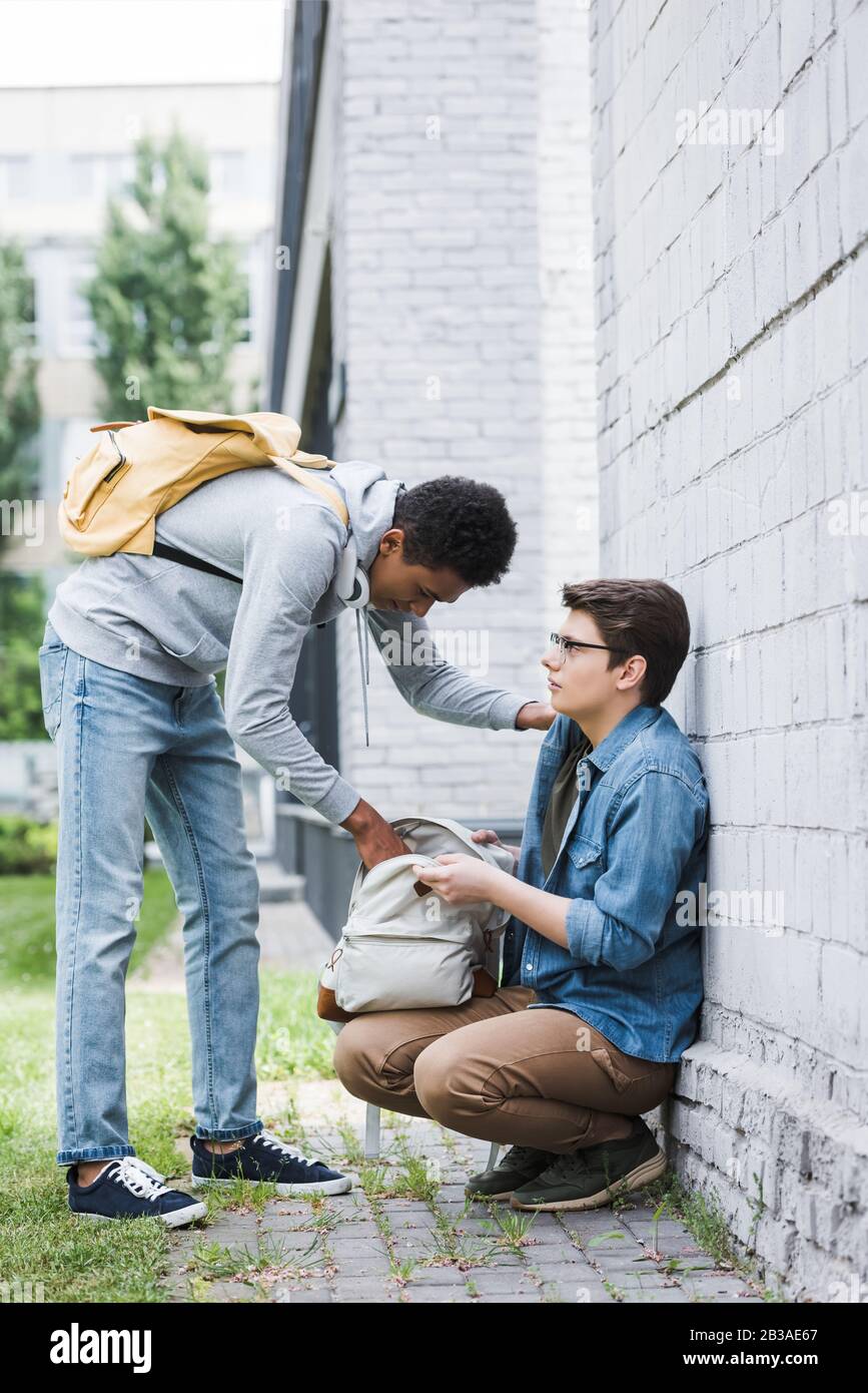 african american boy taking backpack from frightened boy in glasses ...