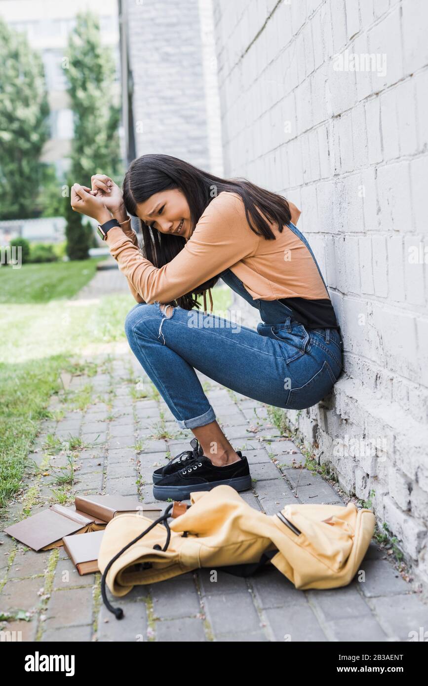 crying brunette teenager in overalls sitting near wall among books ...