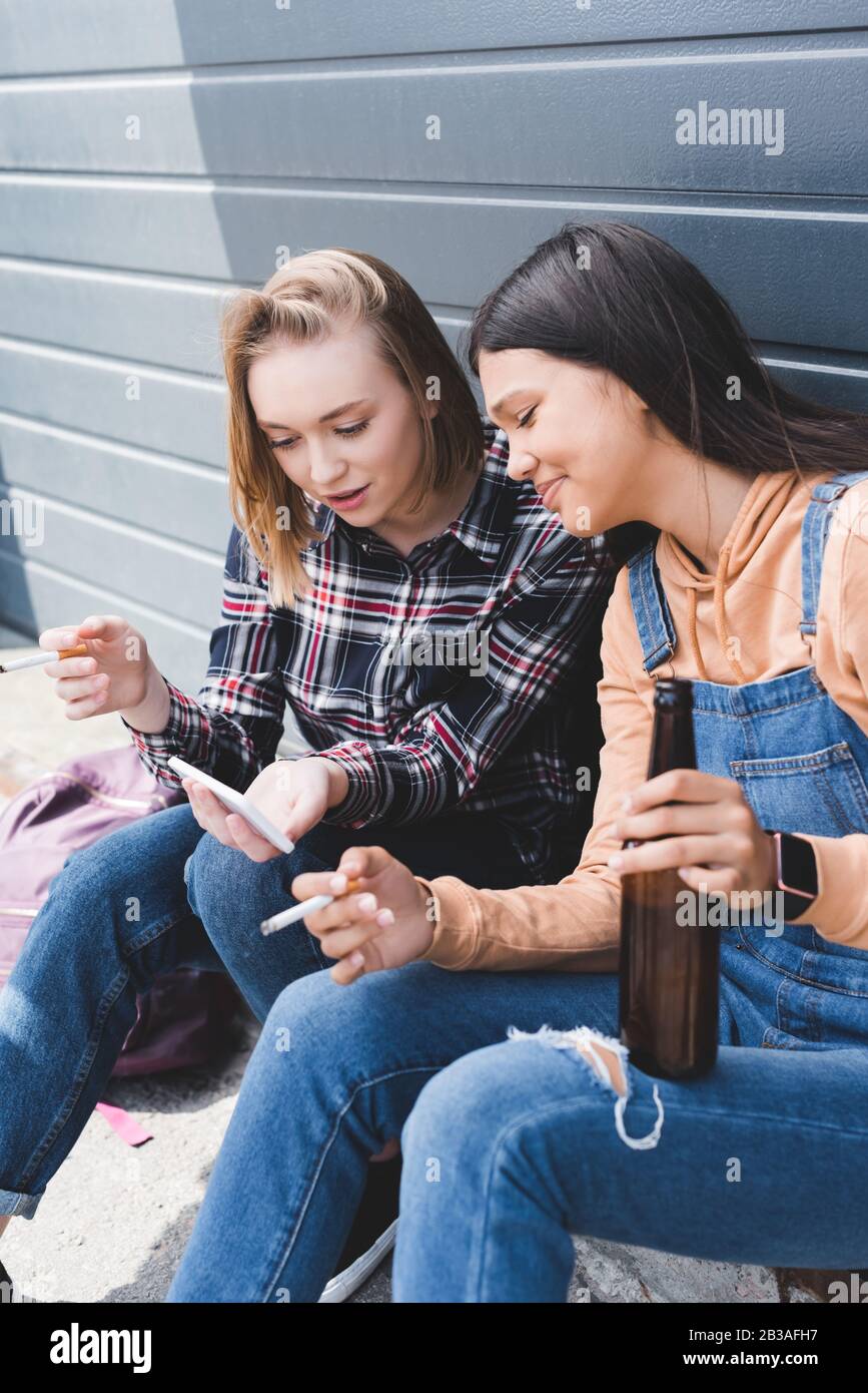 pretty friends smoking cigarettes, holding beer, sitting and looking at ...