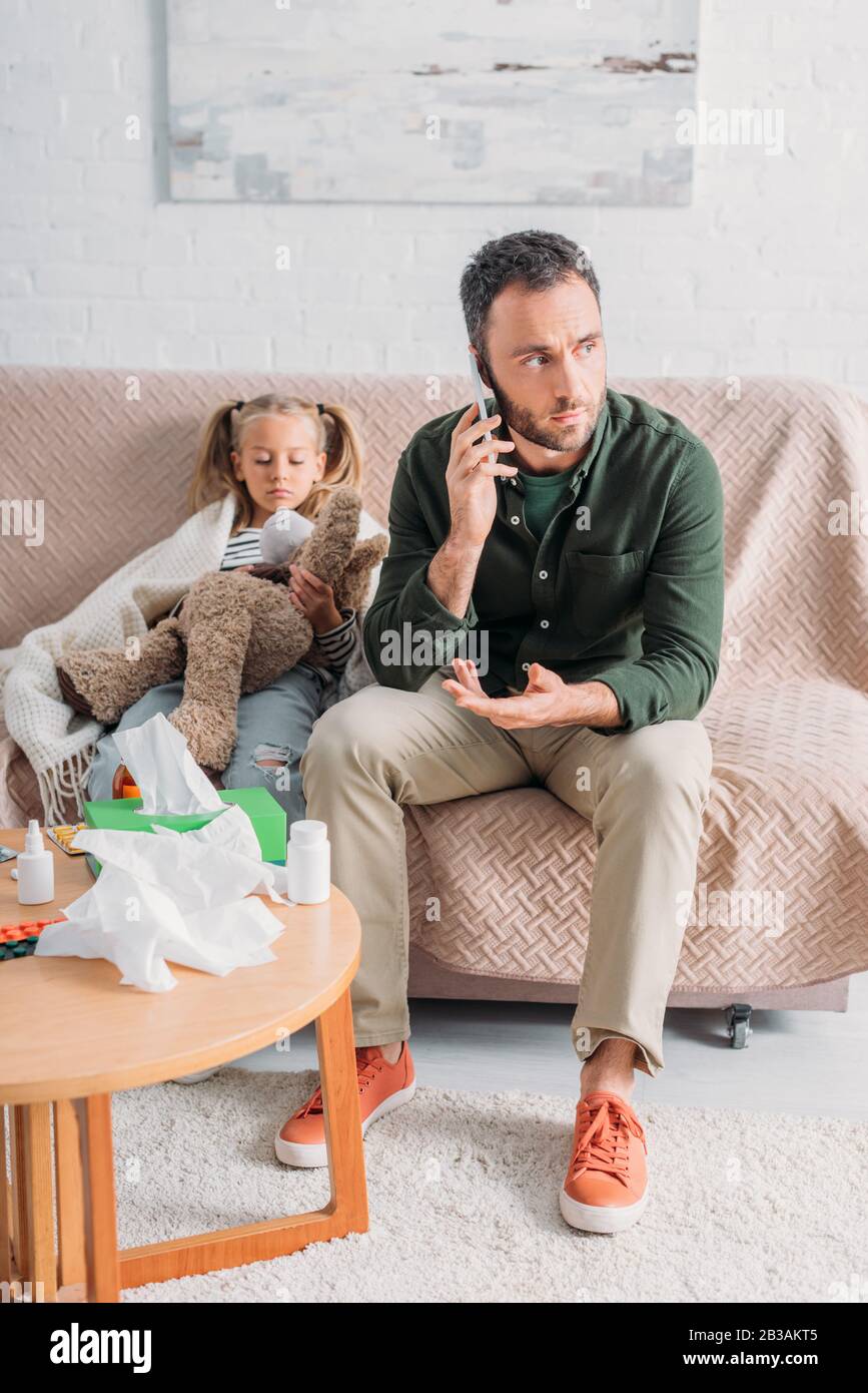 serious father talking on smartphone while sitting near sick daughter ...
