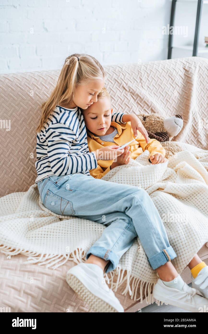 attentive sister looking at thermometer while hugging sick brother ...