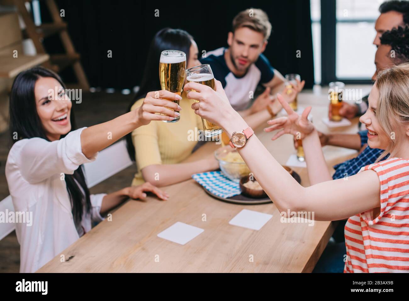 happy young woman clinking glasses of light beer while sitting in pub ...