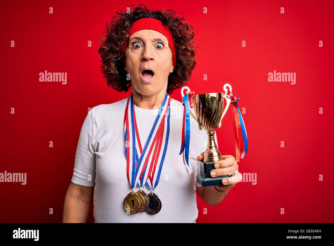 Middle age curly woman winning medals holding trophy over isolated red ...