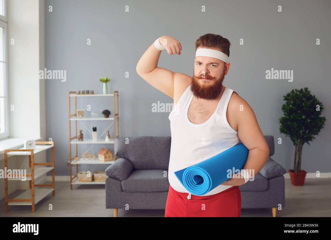Funny fat man in sportswear shows muscles standing in the room at home ...