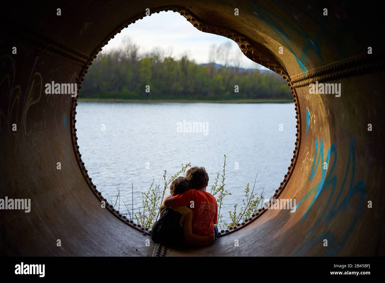 Two young brothers sit together and embrace inside empty rusty drain ...