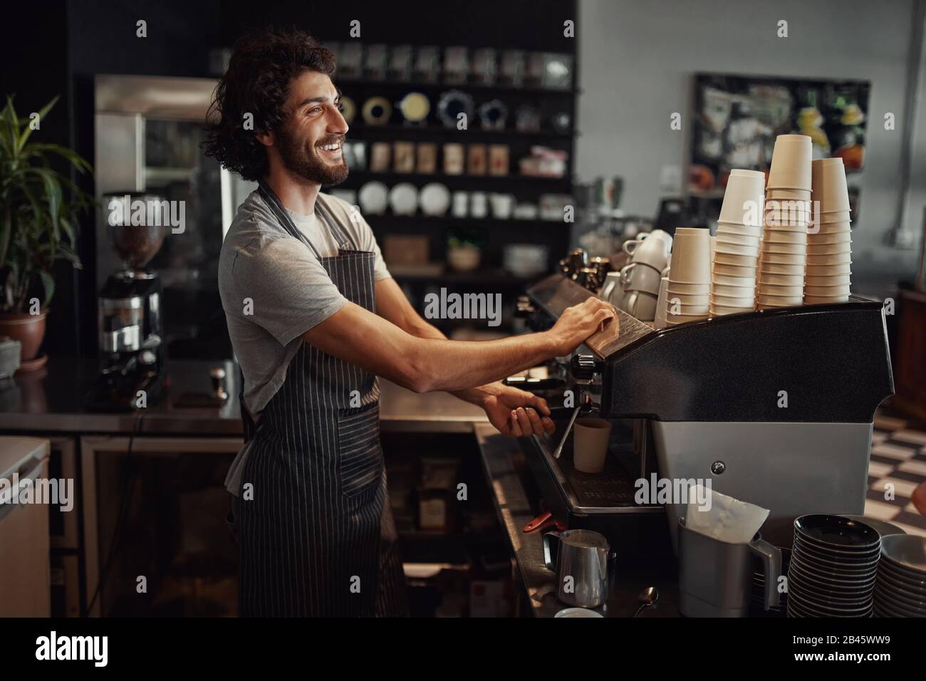 Young cheerful barista wearing black apron while preparing coffee at an automatic machine in a ...