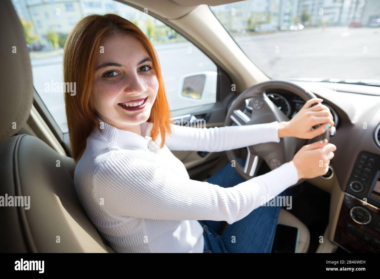 Young redhead woman driver behind a wheel driving a car smiling happily ...