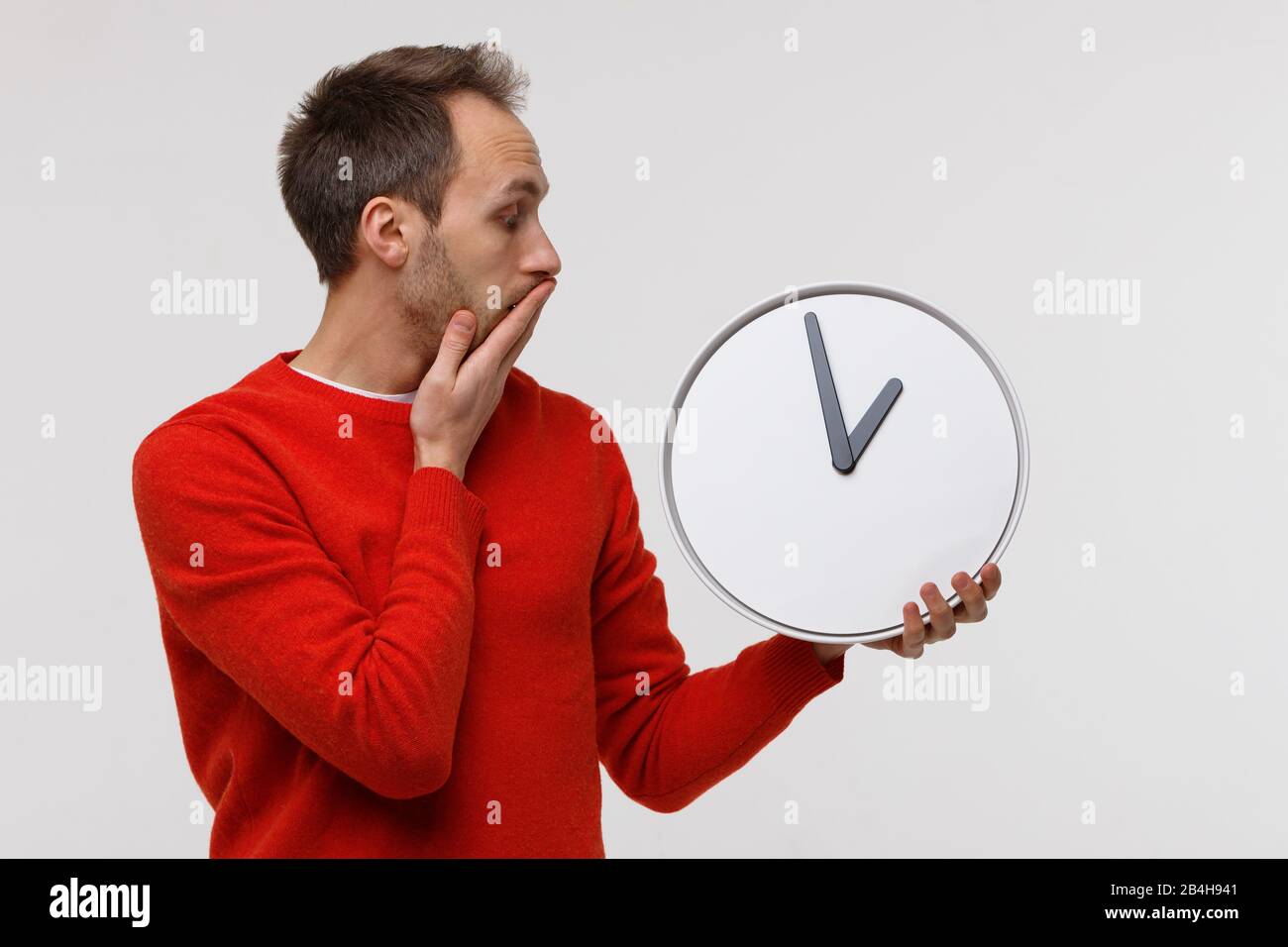 Man in red sweater holding big round clock, in shock covers his mouth ...