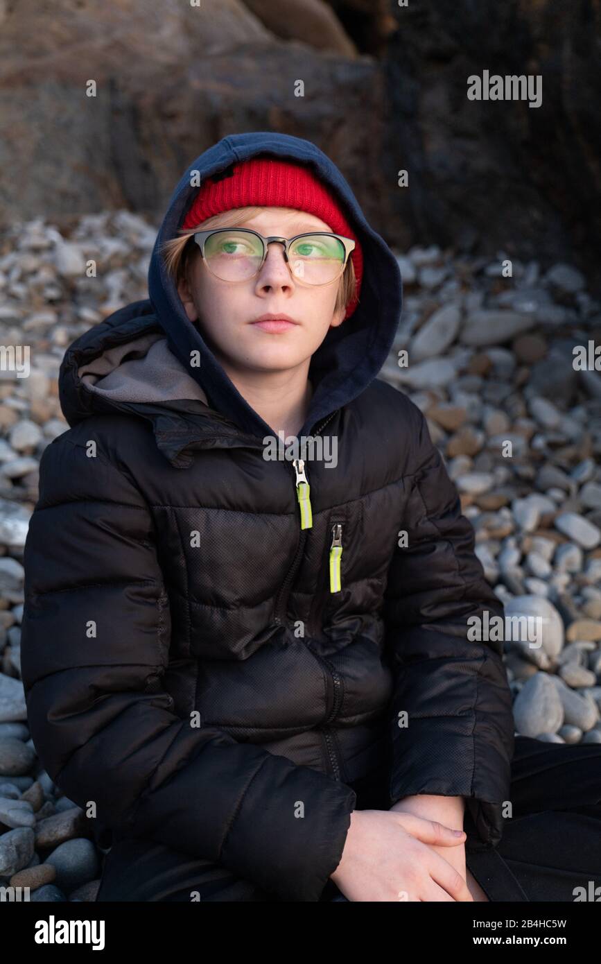Tween boy sitting on beach of stones wearing glasses watching sunset ...