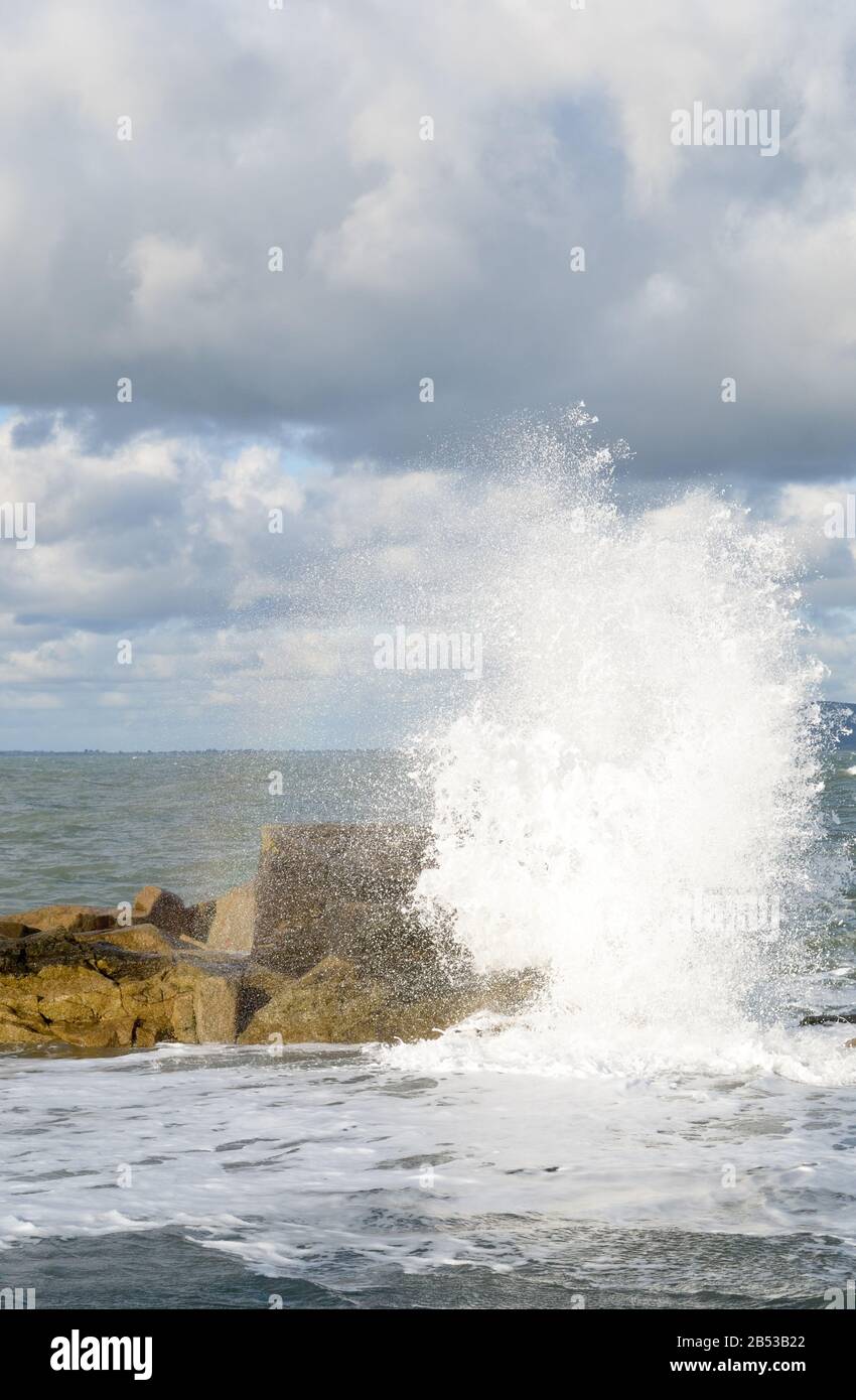 Waves crash onto rocks illuminated by the sun,overcast sky in backround ...