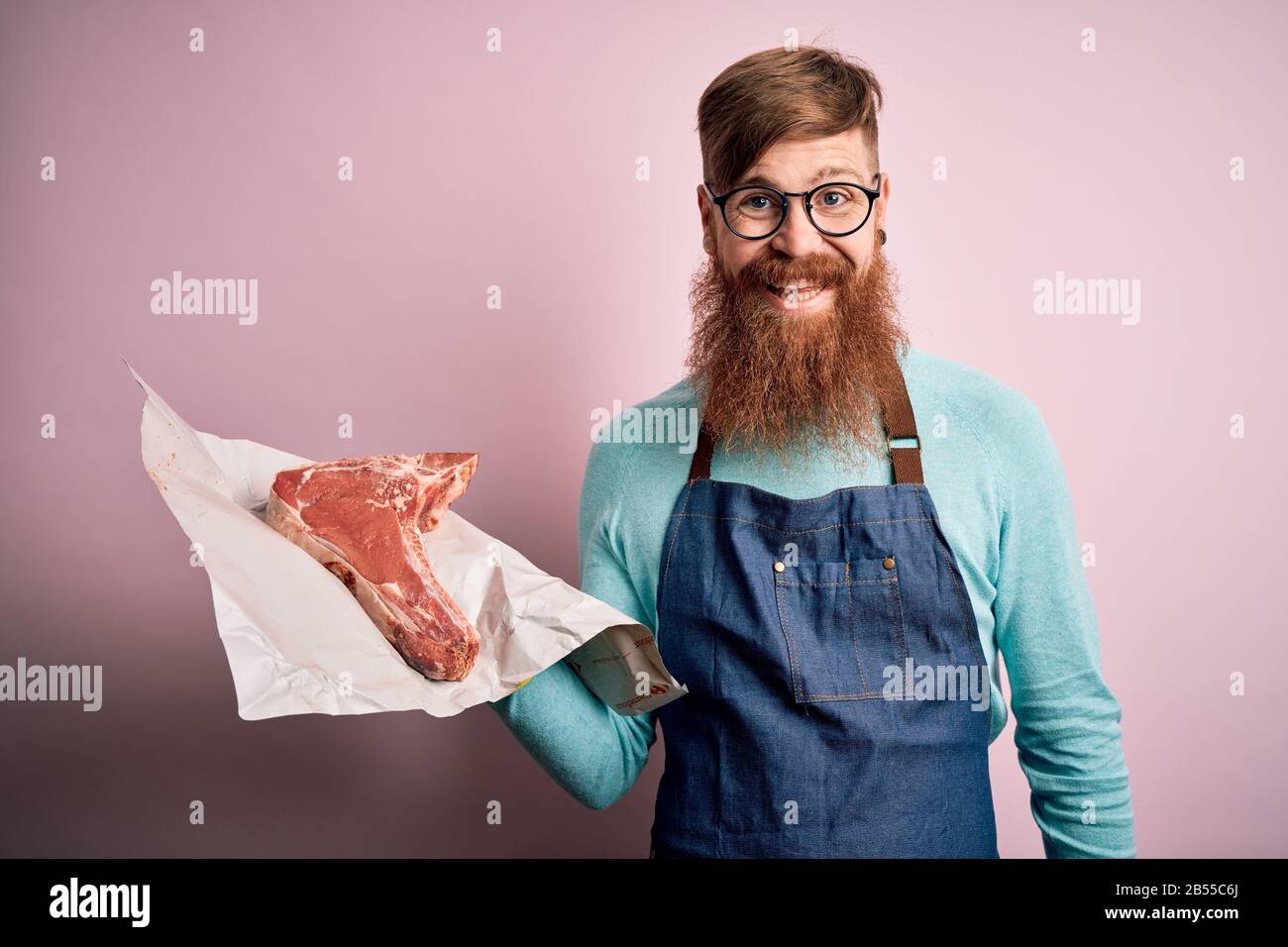 Redhead Irish butcher man with beard holding raw beef steak over pink ...