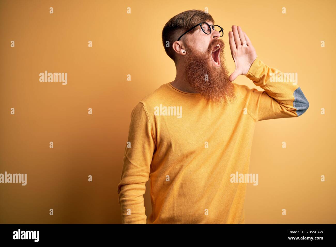 Handsome Irish redhead man with beard wearing glasses over yellow ...