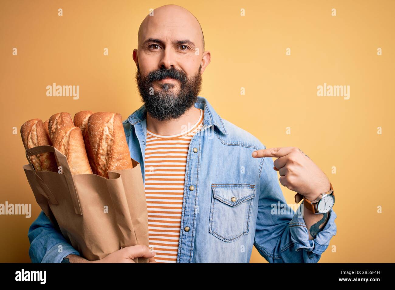 Handsome bald man with beard holding paper bag with bread over yellow ...