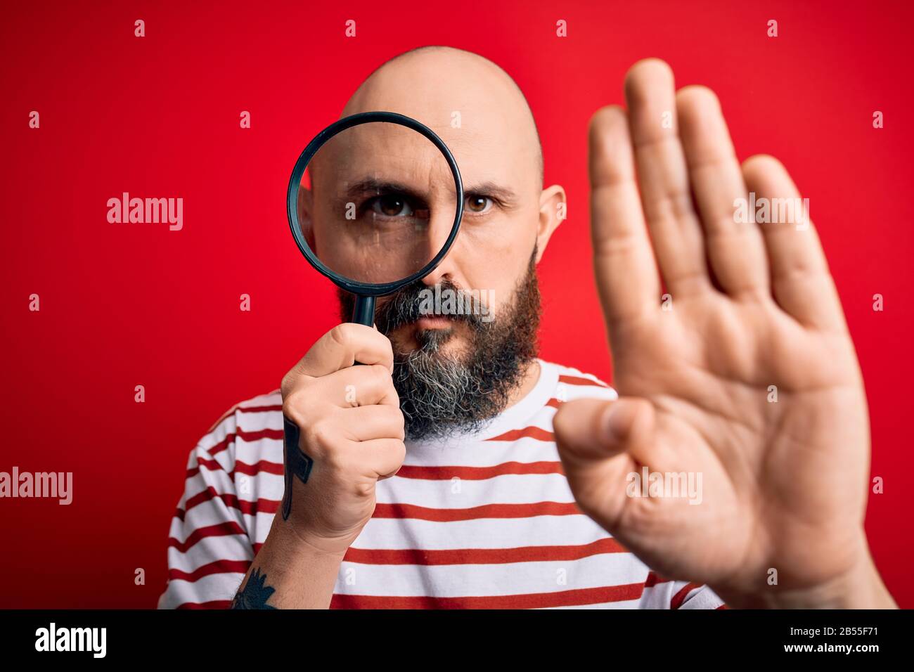 Handsome detective bald man with beard using magnifying glass over red ...