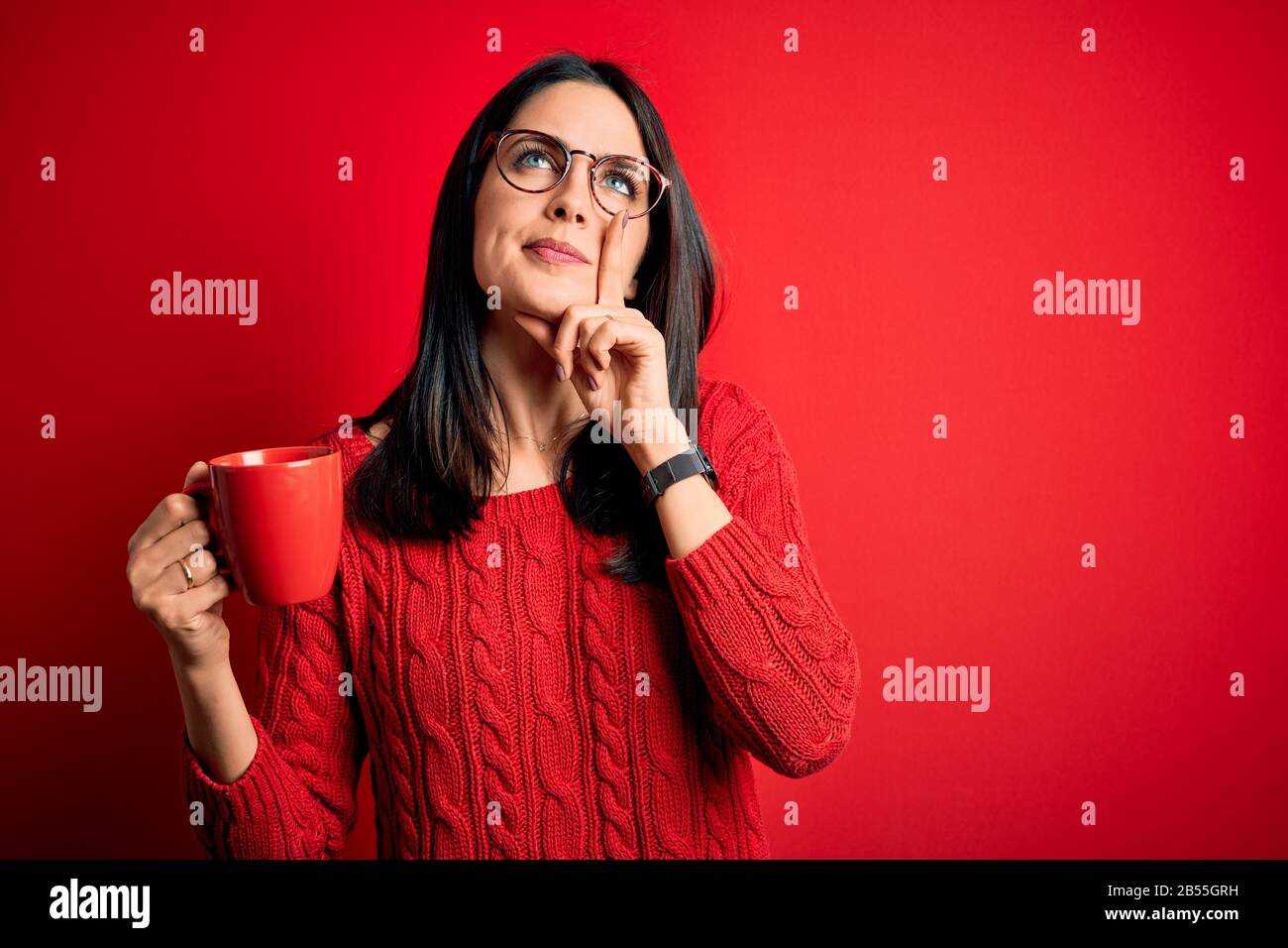 Young brunette woman with blue eyes wearing glasses and drinking a cup of coffee serious face ...