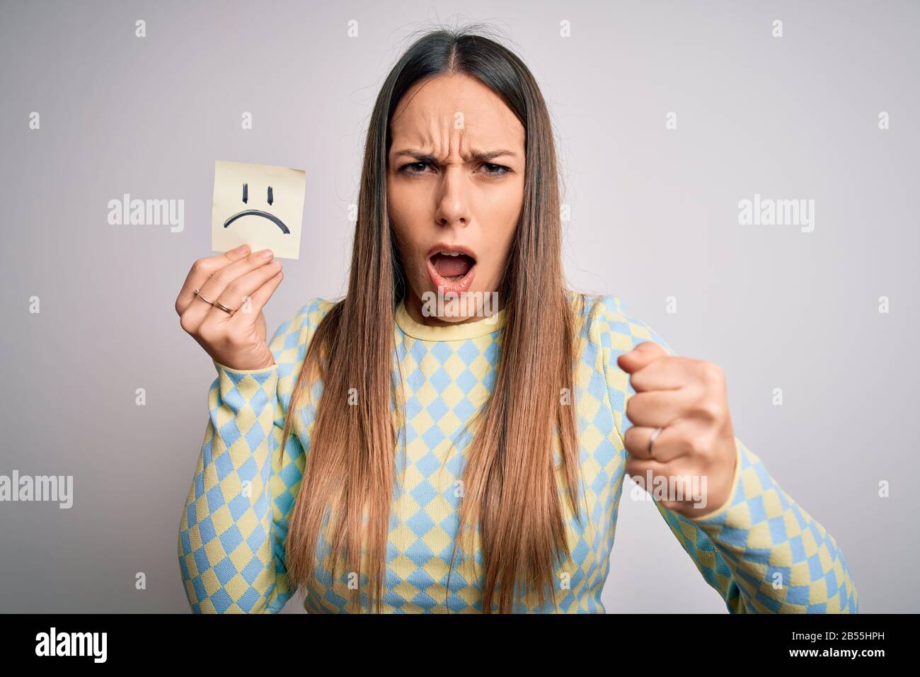 Young blonde woman holding sticky paper note with sad face emoticon ...