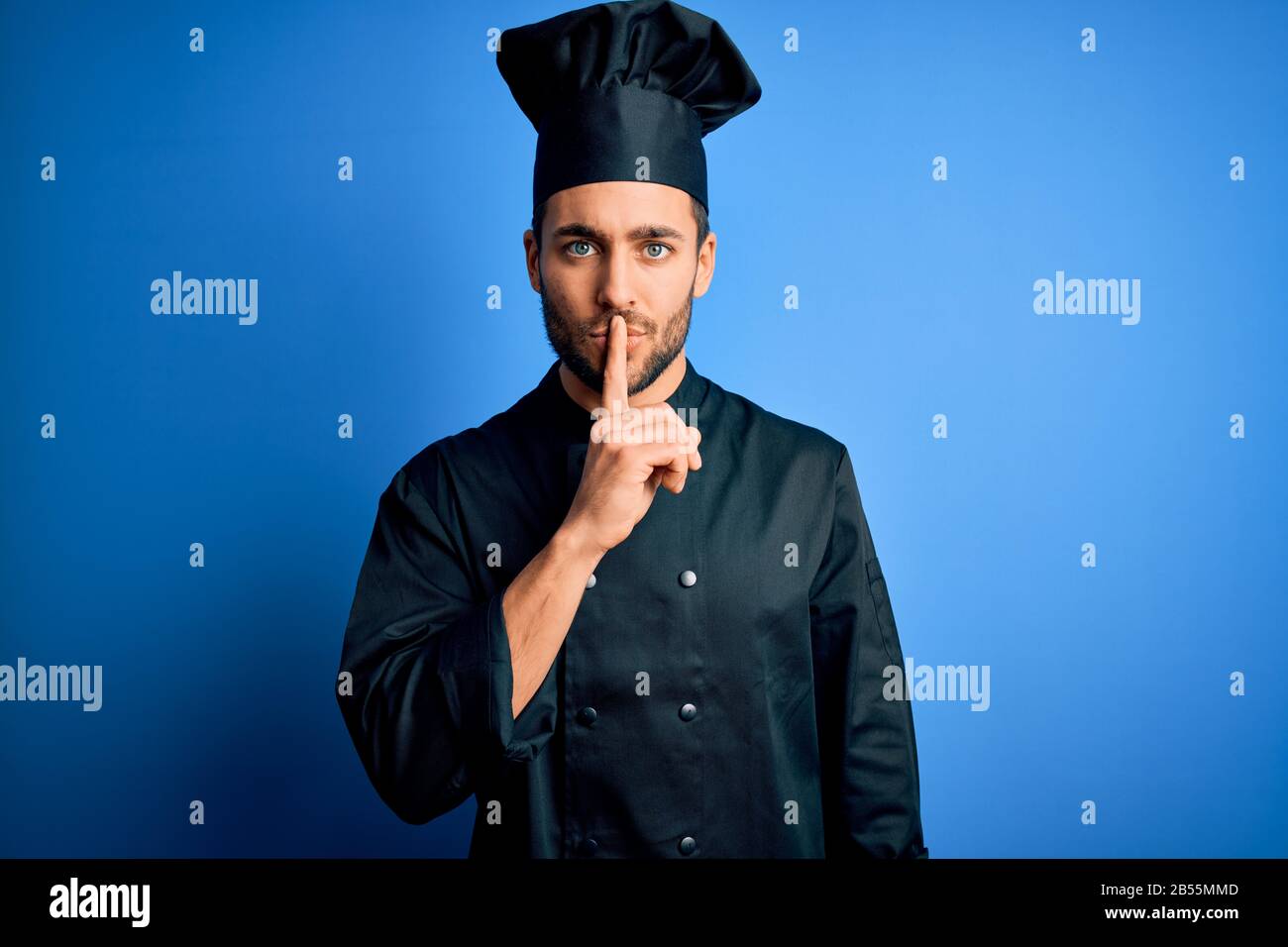 Young handsome chef man with beard wearing cooker uniform and hat over ...