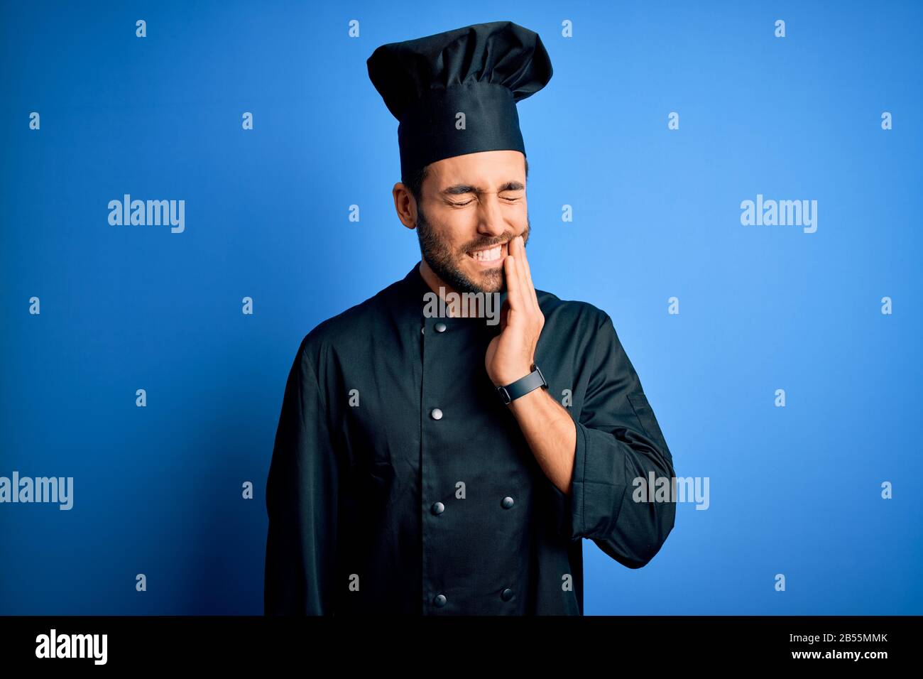 Young handsome chef man with beard wearing cooker uniform and hat over ...