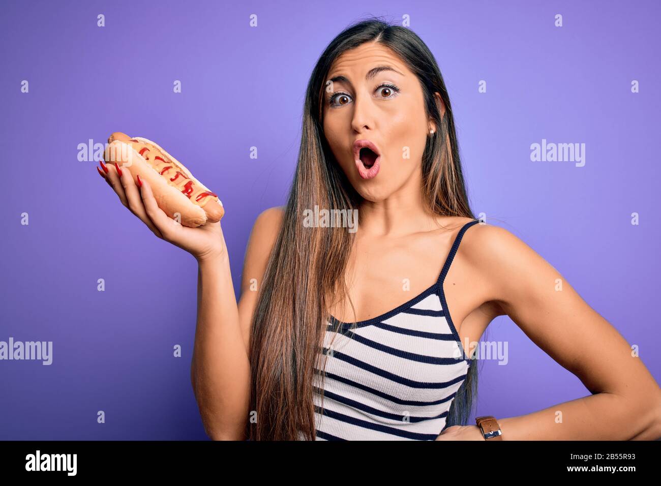 Young woman eating hotdog with ketchup and mustard over purple ...