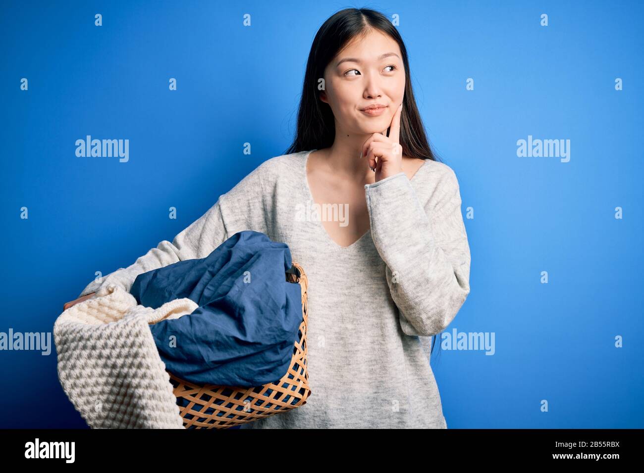 Young asian woman holding wicker laundry basket doing domestic chores serious face thinking ...