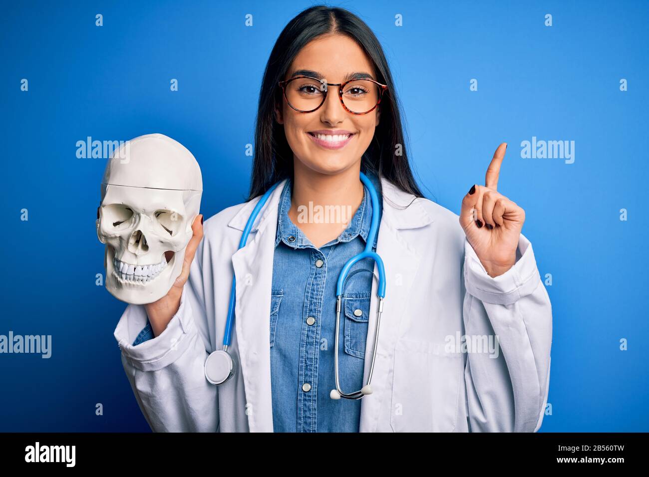 Young beautiful brunette doctor woman wearing coat holding human skull ...