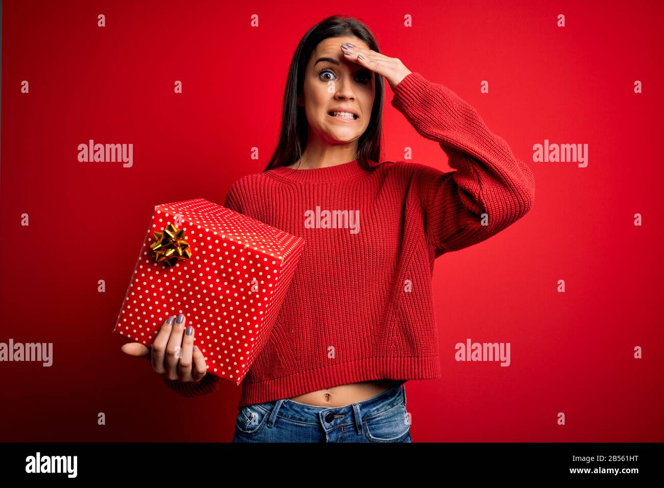 Young beautiful brunette woman holding birthday gift over isolated red ...