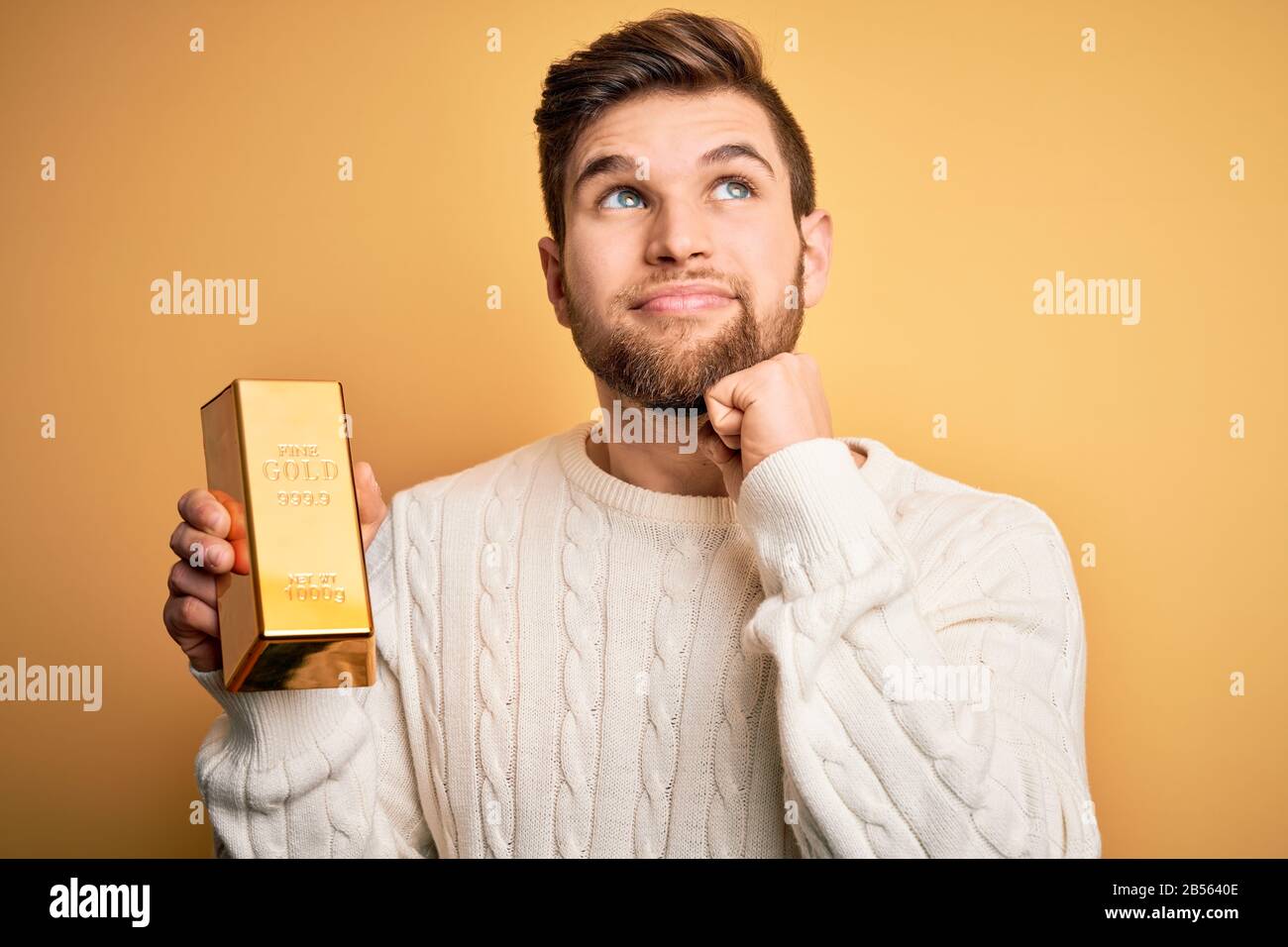 Young blond man with beard and blue eyes holding gold ingot over ...