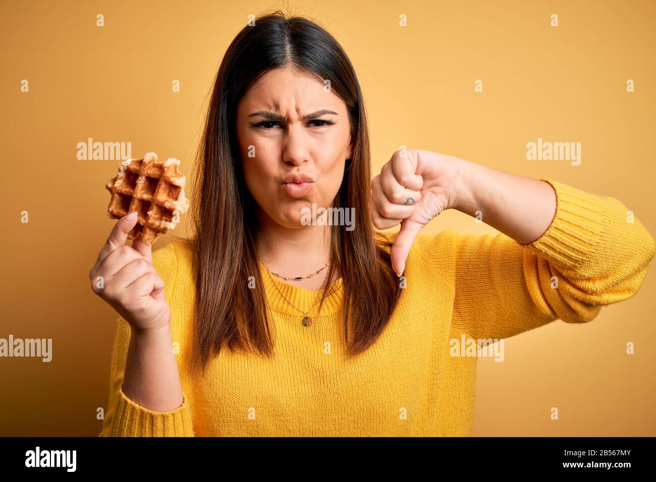 Young beautiful woman eating sweet waffle pastry over yellow background ...