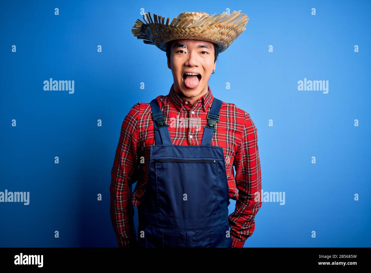 Young handsome chinese farmer man wearing apron and straw hat over blue ...