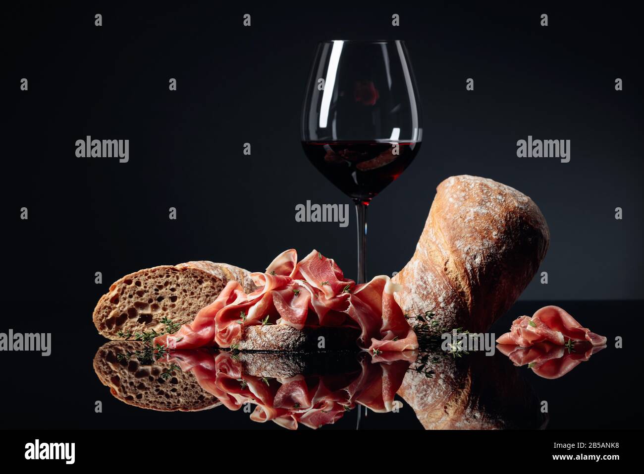 Prosciutto with ciabatta, red wine and thyme on a black reflective ...