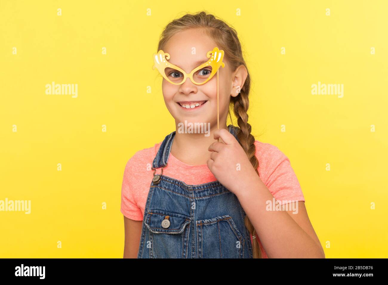 Portrait of positive carefree little girl with braid in denim overalls ...