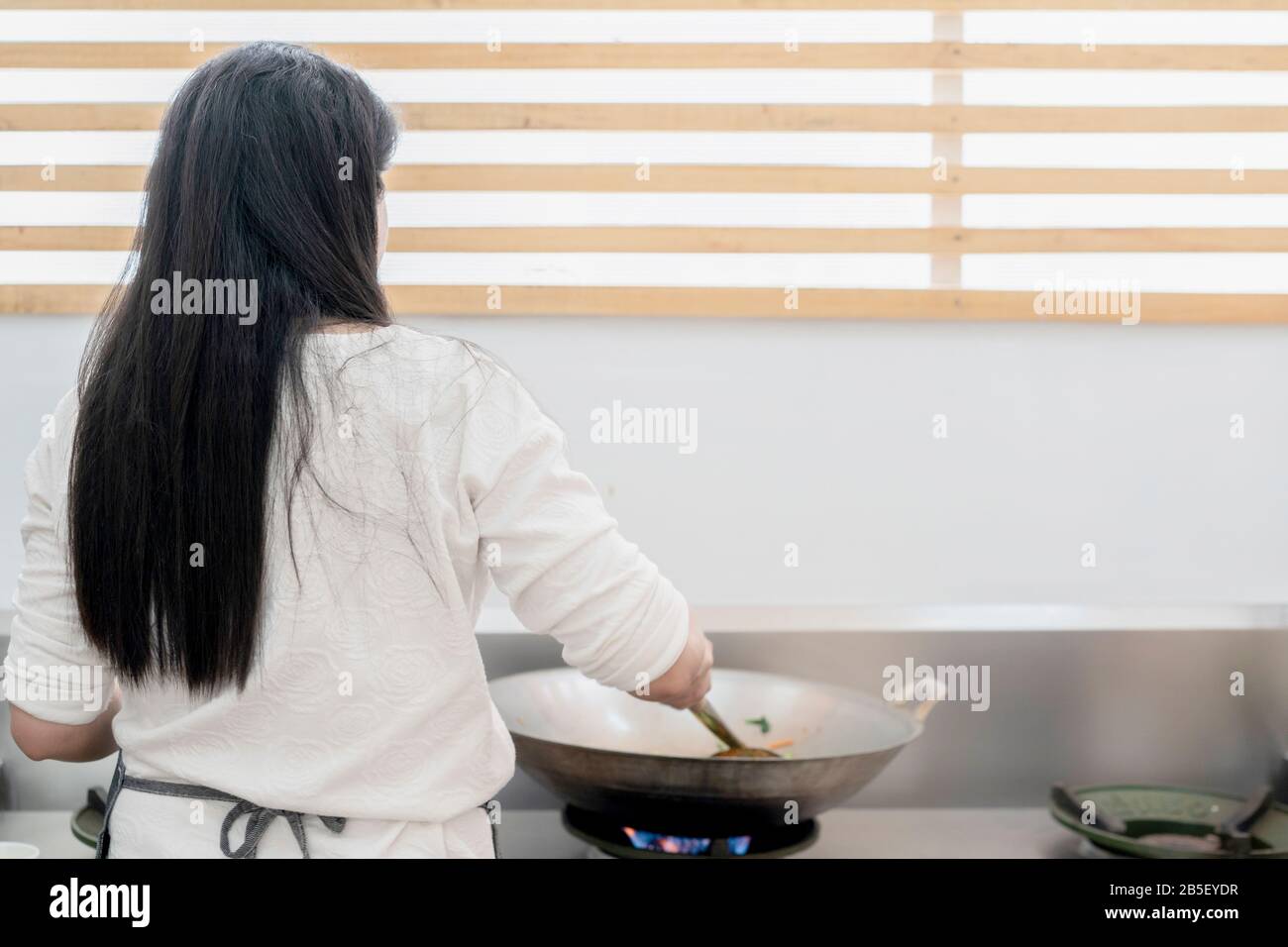 Asian female with long black hair cooks food in steel pan on stove with ...