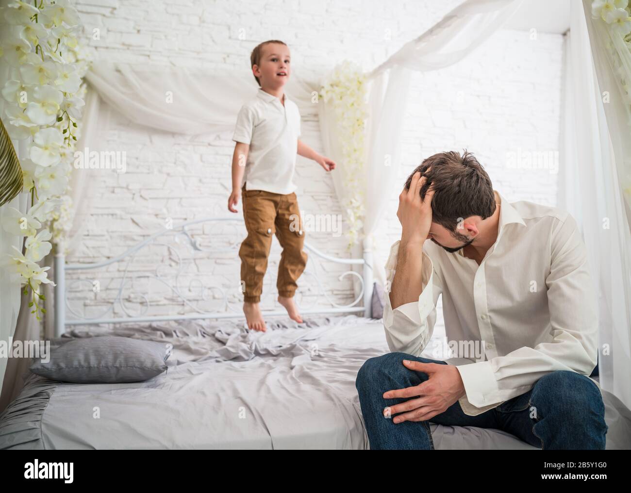 Tired young father is sitting on the edge of a large and beautiful bed ...