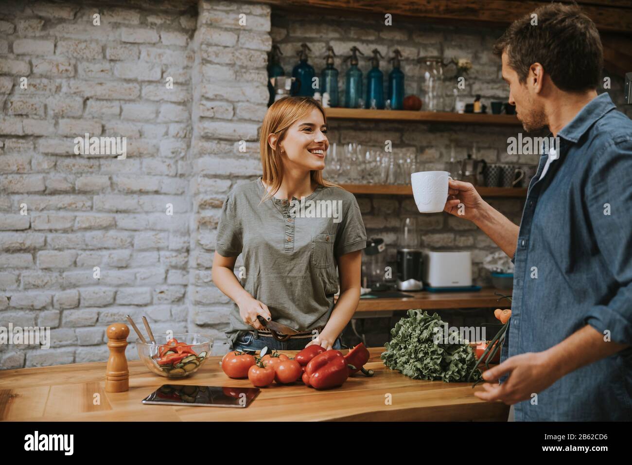 Young woman cooking while man drinking coffee in the rustic kitchen ...