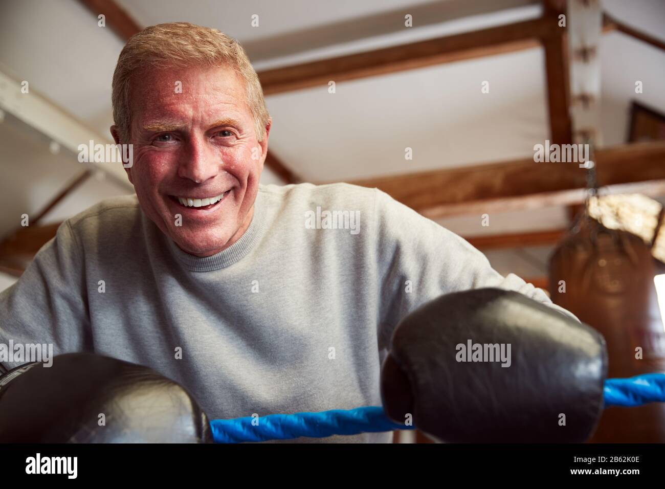 Smiling Senior Male Boxer In Gym Wearing Boxing Gloves Leaning On Ropes ...