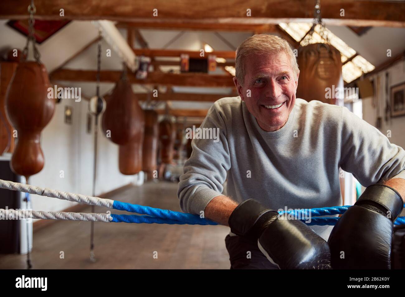 Smiling Senior Male Boxer In Gym Wearing Boxing Gloves Leaning On Ropes ...