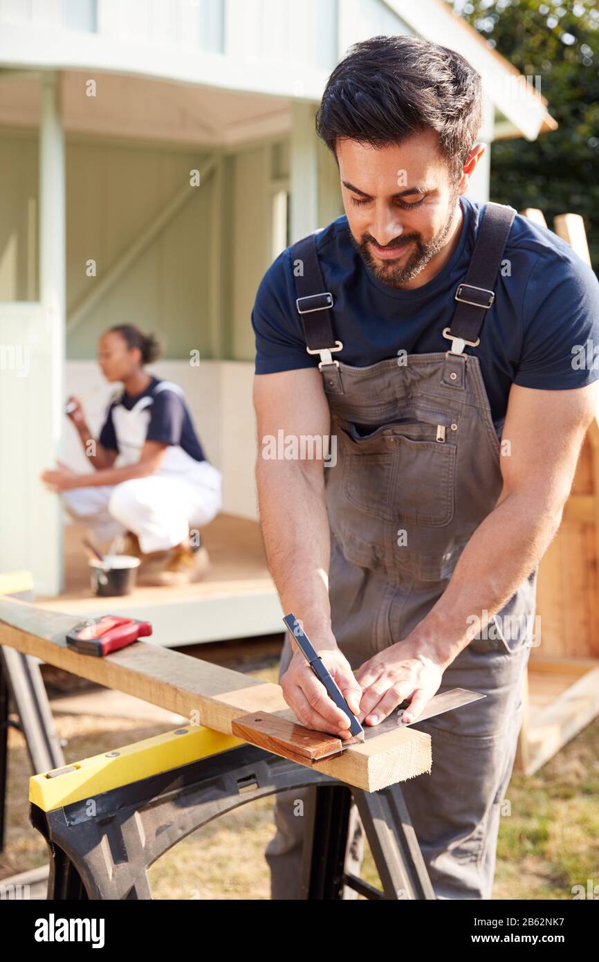 Male Carpenter With Female Apprentice Measuring Wood To Build Outdoor ...