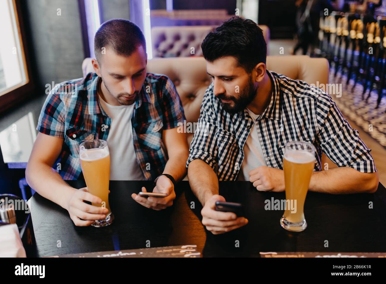 Two happy young men in casual wear drinking beer in pub while one of ...