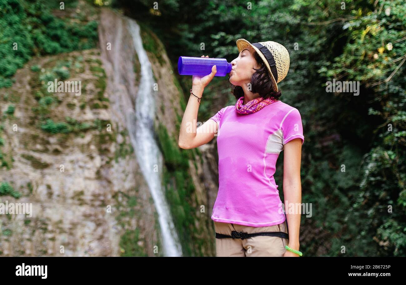 Healthy hiker girl drinking water in bottle in front of waterfall ...
