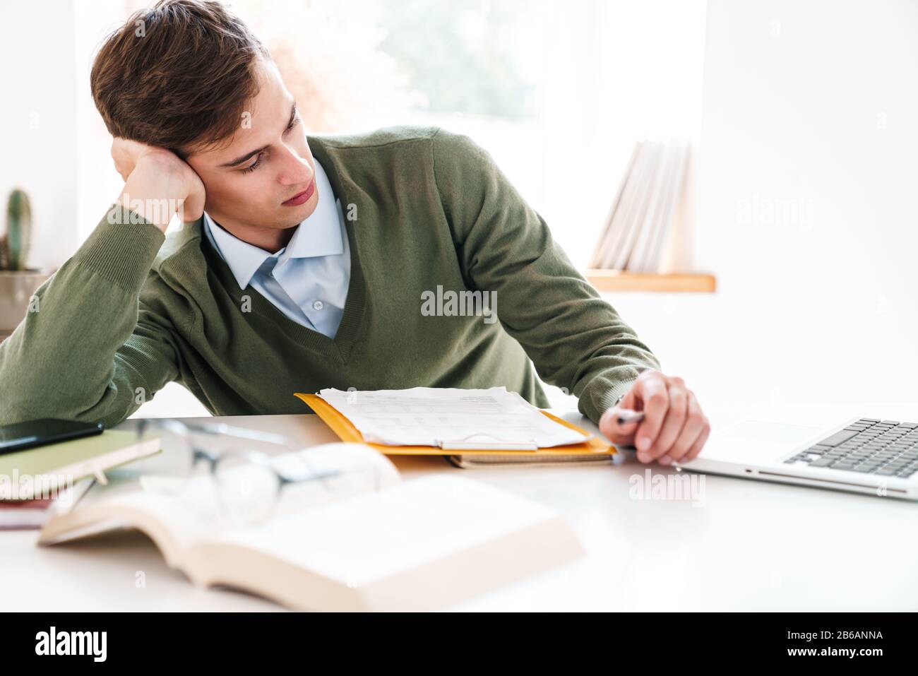 Image of bored tired young guy student sit at the table indoors doing ...
