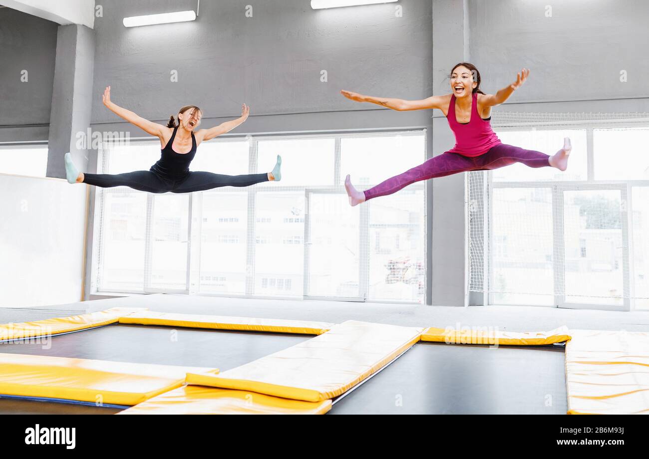 Two young woman friends jumping on a trampoline and doing split indoors ...