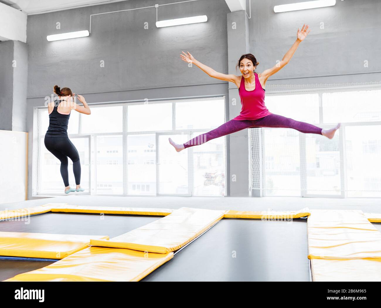 Two young woman friends jumping on a trampoline and doing split indoors ...