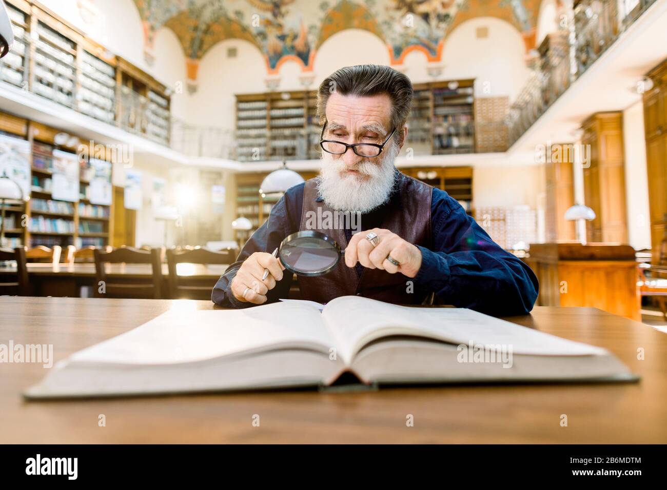 Old man scientist, library worker, reading a book in a library, looking ...