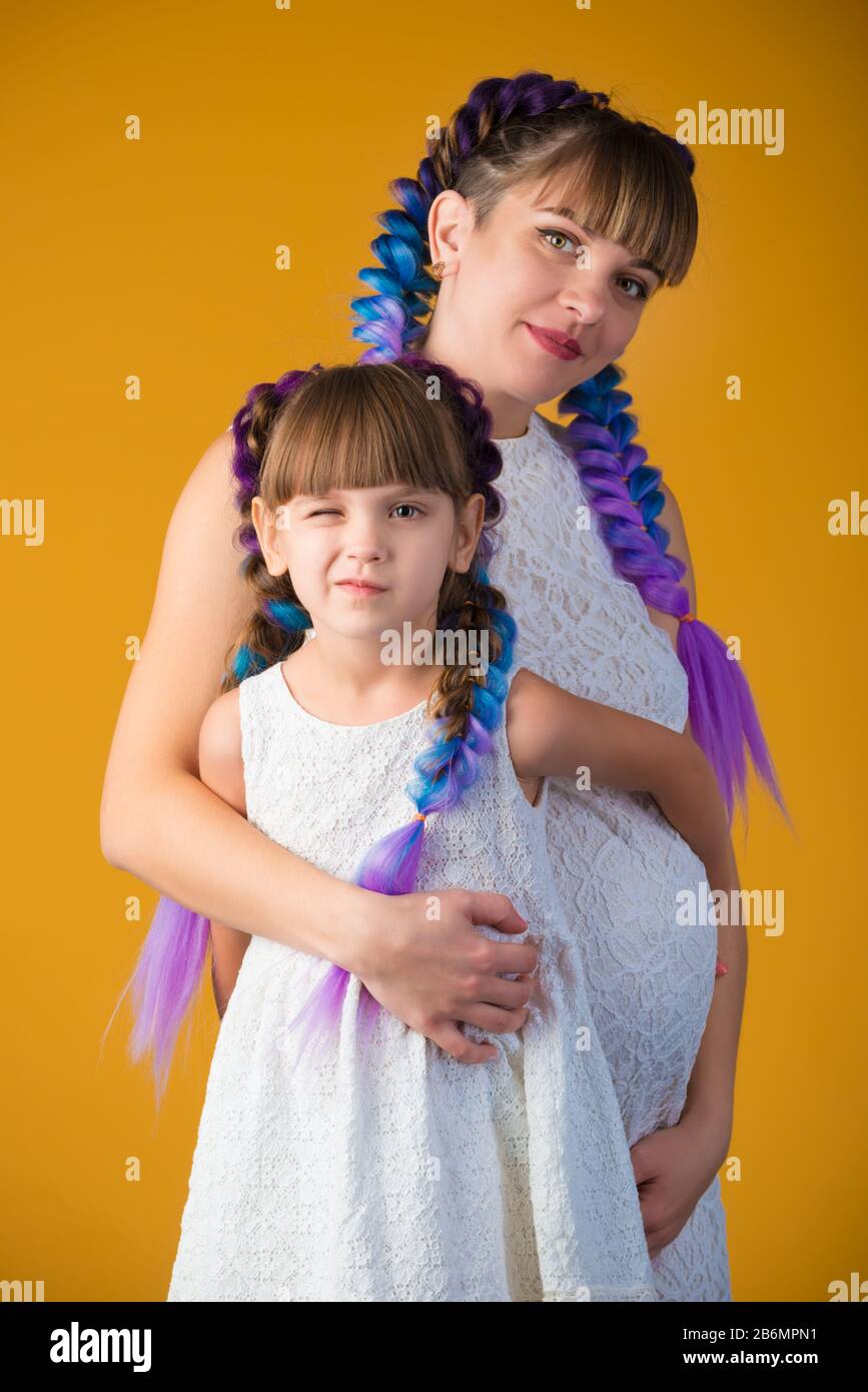 Funny positive mom and daughter in identical white sundresses with colored braids posing in ...