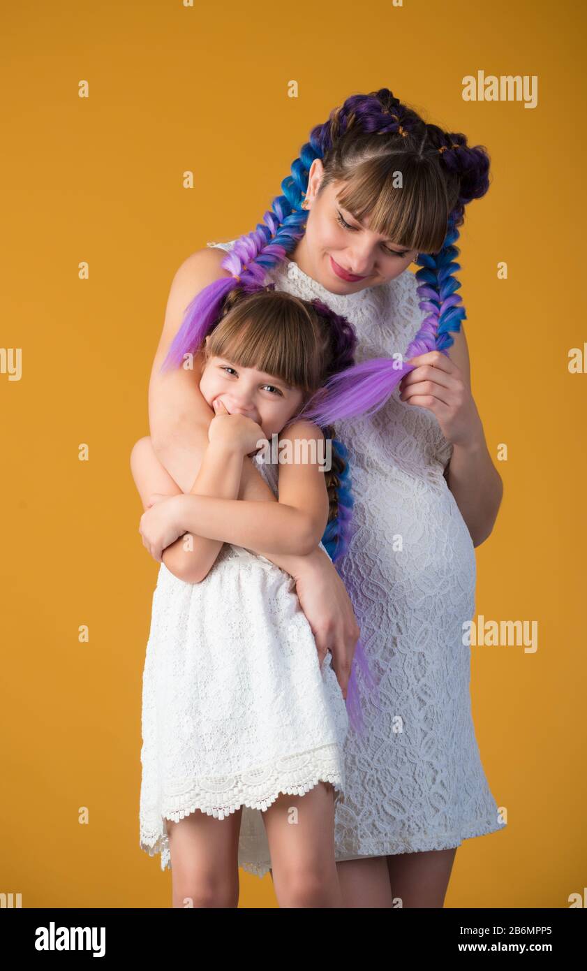 Funny positive mom and daughter in identical white sundresses with colored braids posing in ...