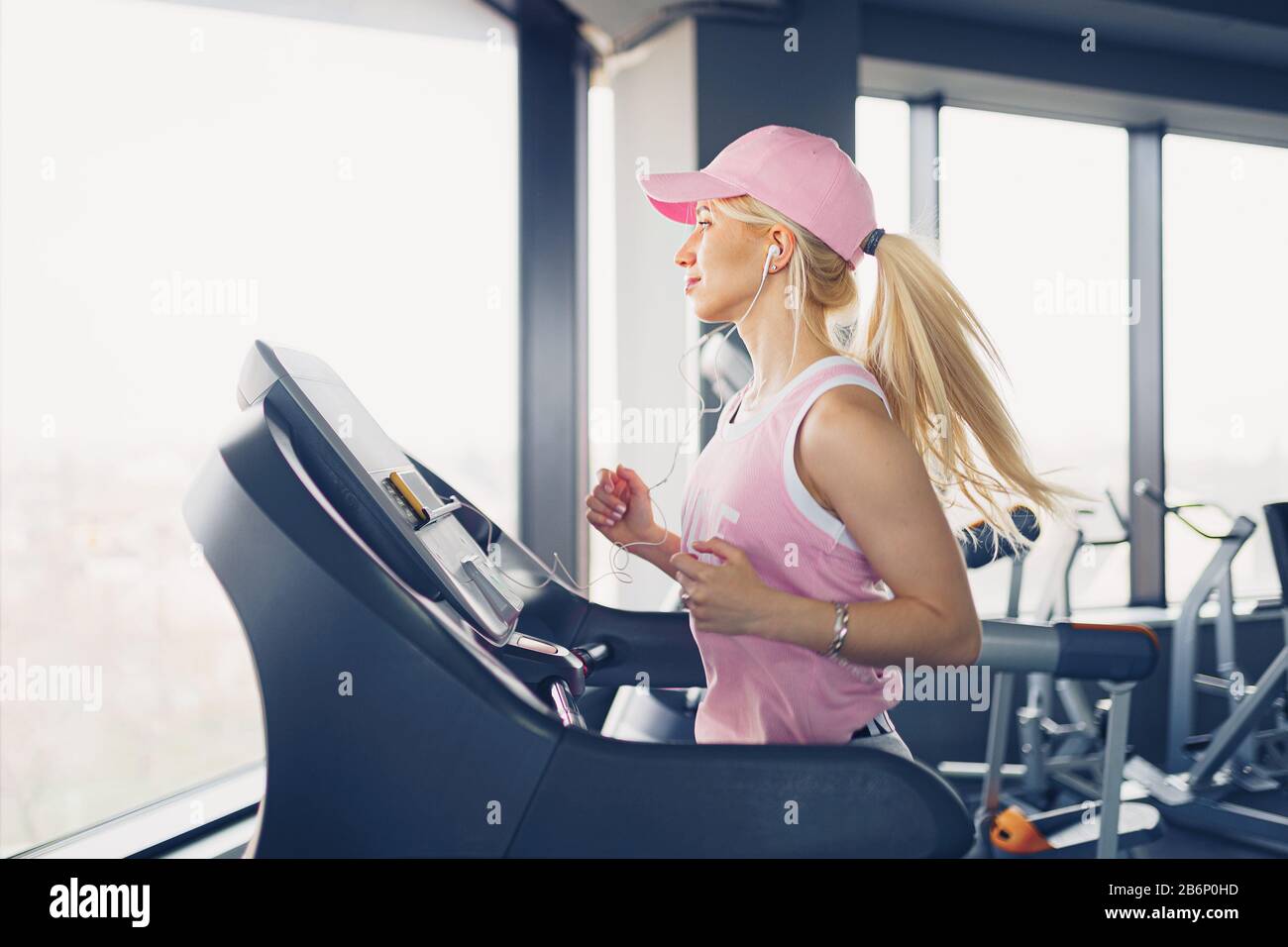 Side view of sporty blonde woman in pink cap exercising on treadmill in ...